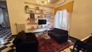 Uma sala de estar com um sofá preto, uma mesa de centro, uma estante, uma televisão e um tapete estampado.