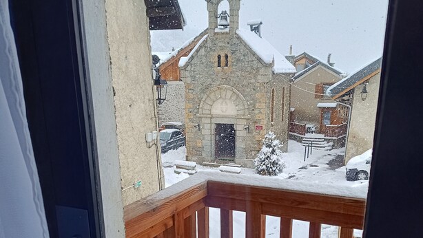 Vista de uma varanda com guarda-corpo de madeira, com vista para uma igreja de pedra e um campanário, rodeada por edifícios cobertos de neve.