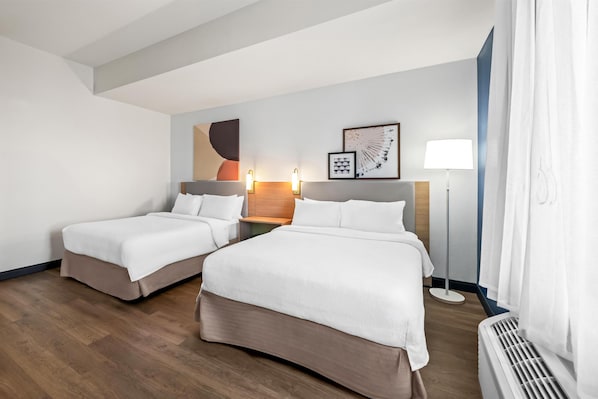Quarto de hotel com duas camas, uma janela grande com cortinas, um abajur e obras de arte na parede.