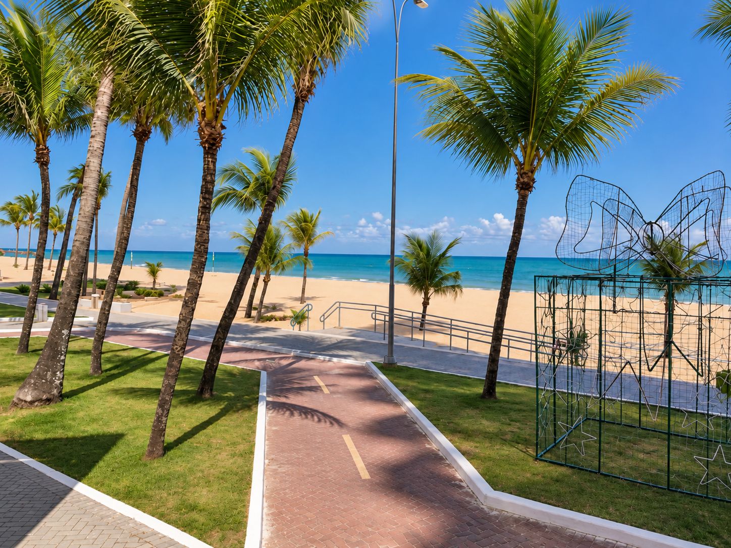 Uma trilha pavimentada leva a uma praia com palmeiras e um céu azul límpido.