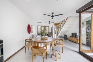 Uma sala de estar ampla com uma mesa de madeira redonda rodeada por cadeiras de vime, uma escada que leva a um andar superior e um grande vão de vidro que dá para o exterior.