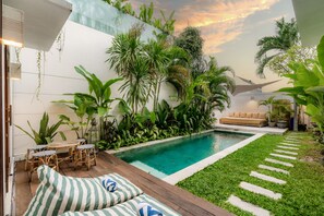 Uma área à beira da piscina com espreguiçadeira, mesa pequena e diversas plantas tropicais.