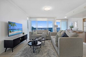 Uma sala de estar moderna com uma grande televisão de tela plana, dois sofás, uma mesa de centro e vista para o oceano através de grandes janelas.