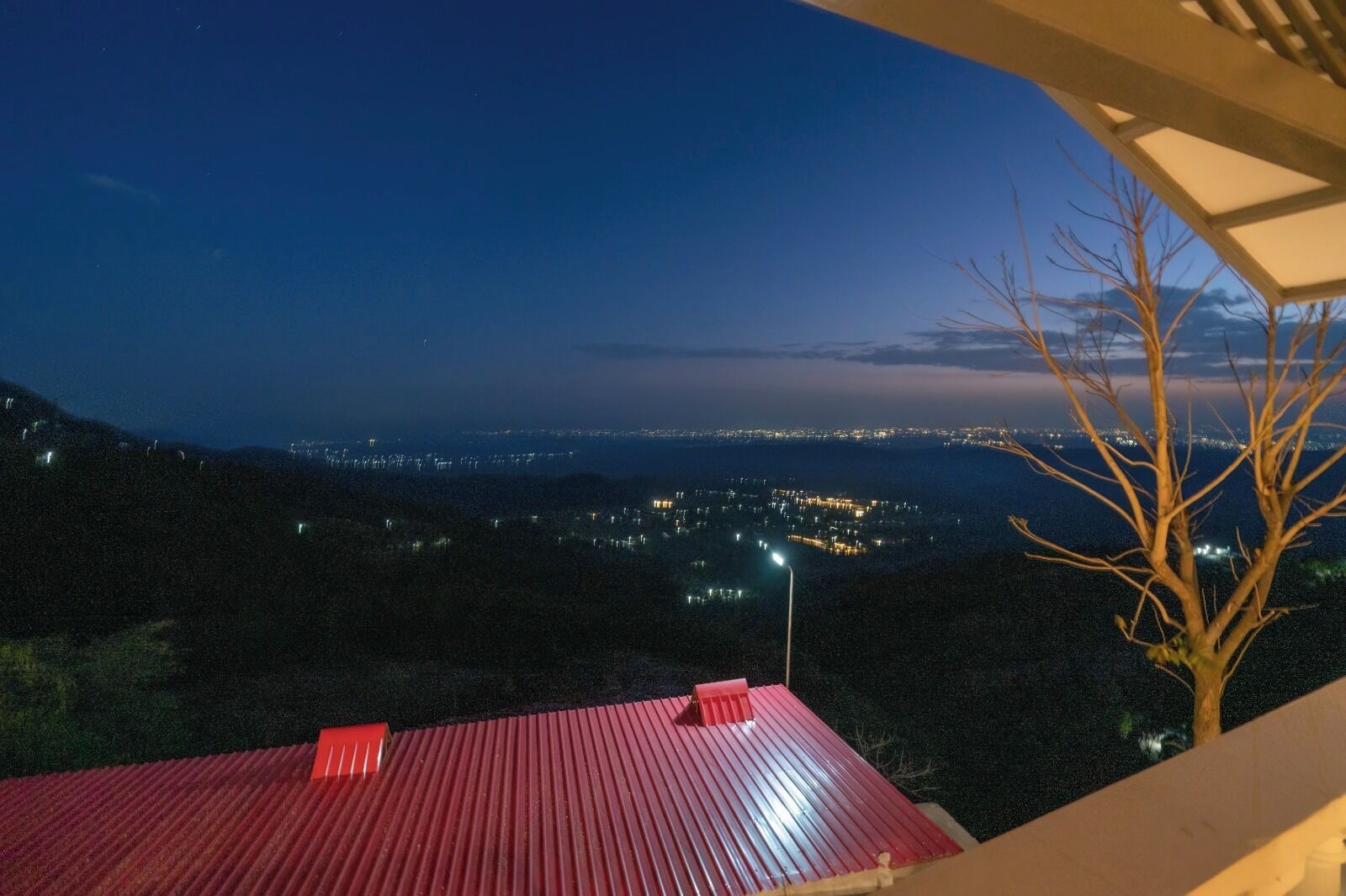 Vista noturna de uma varanda com telhado vermelho, com vista para uma paisagem urbana iluminada e colinas ao longe.
