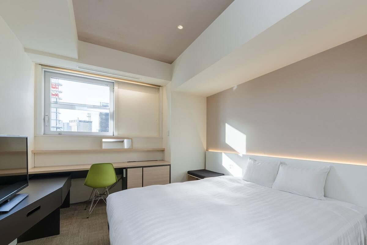 Quarto de hotel moderno com uma cama grande, uma escrivaninha, uma cadeira e uma janela com vista para a cidade.
