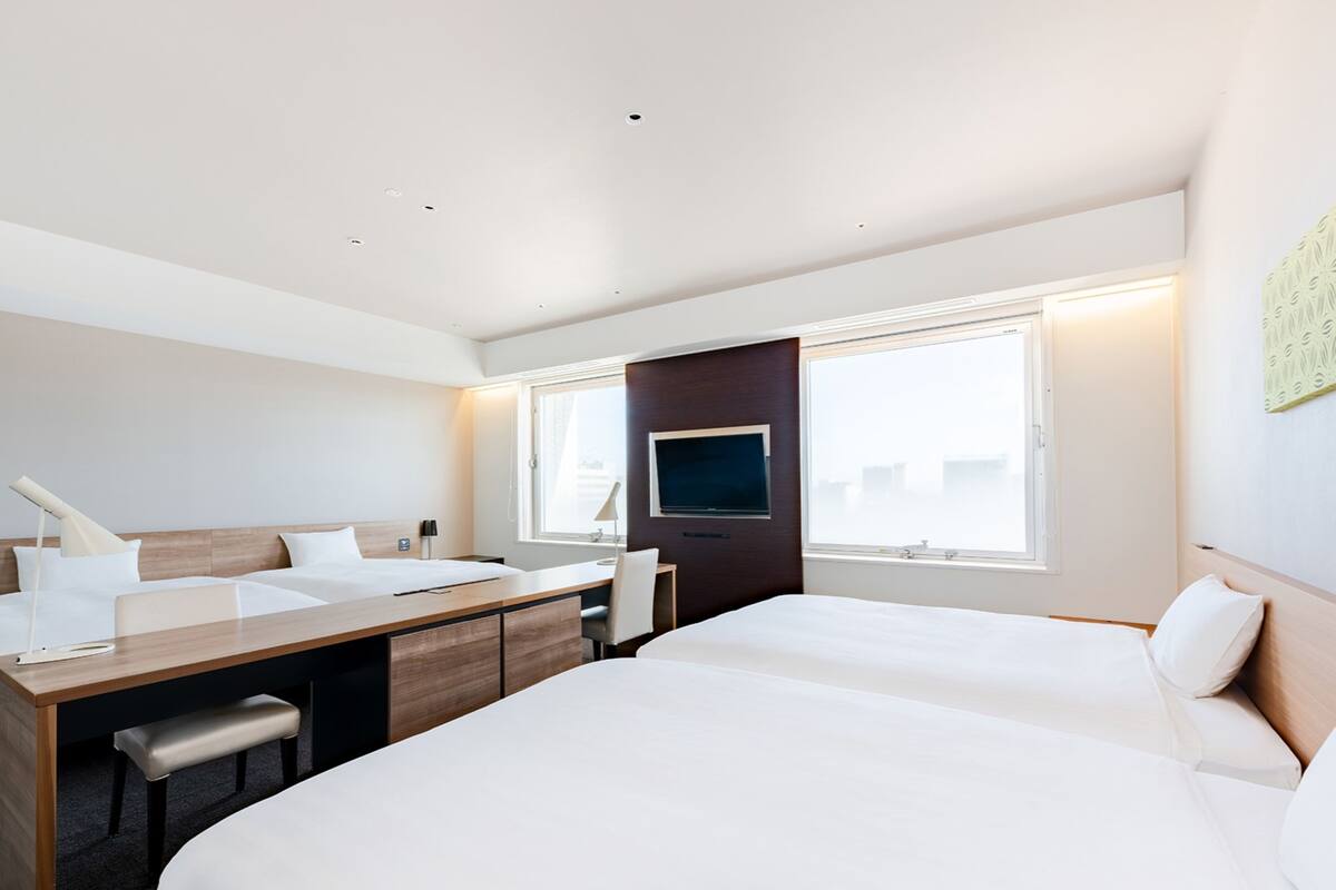 Quarto de hotel moderno com uma cama grande, uma escrivaninha com cadeira, televisão e janela com vista para a cidade.