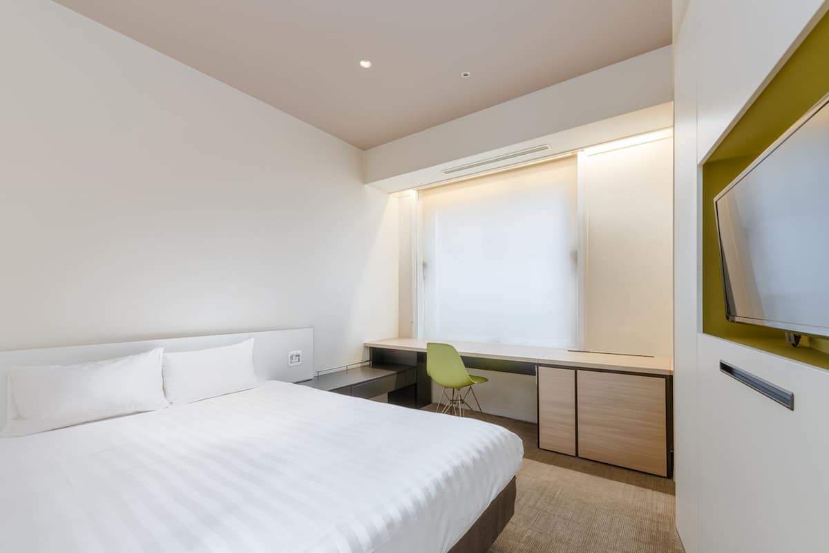 Quarto de hotel moderno com uma cama grande, escrivaninha, cadeira e televisão na parede.