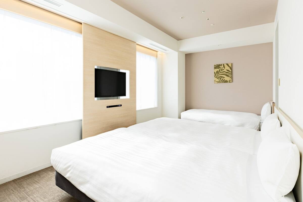 Quarto de hotel com duas camas, uma TV de tela plana fixada em um painel de madeira e uma obra de arte com padrão de folhas na parede.