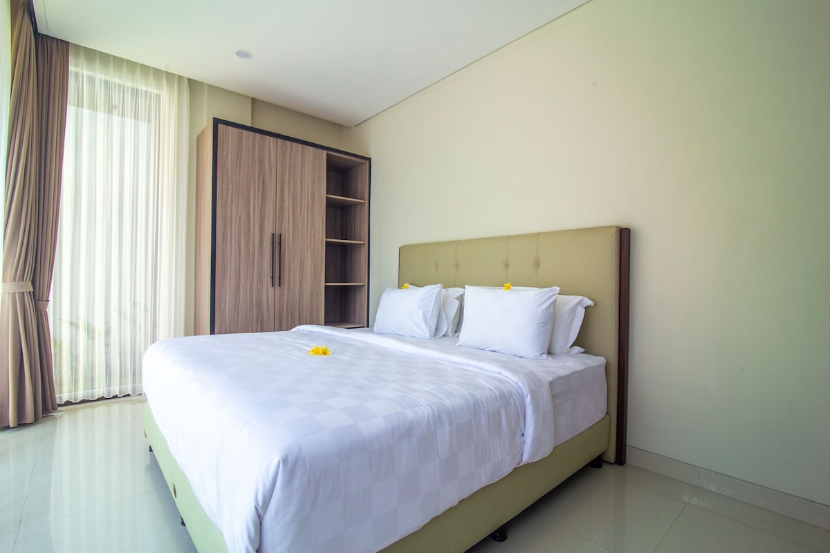 Uma cama bem arrumada com lençóis e almofadas brancas, um guarda-roupa de madeira e uma janela grande com cortinas leves.