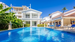 Piscina com água azul cristalina, rodeada por edifícios brancos, palmeiras e espreguiçadeiras.