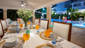 Mesa de jantar posta para seis pessoas, com bebidas azuis e laranjas, pratos brancos e um arranjo floral.