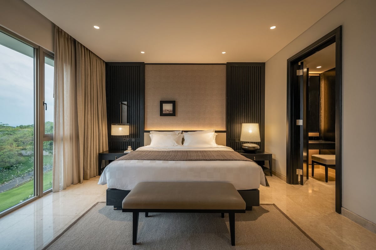 Quarto de hotel moderno com uma cama grande, mesas de cabeceira, um banco e vista para o exterior.