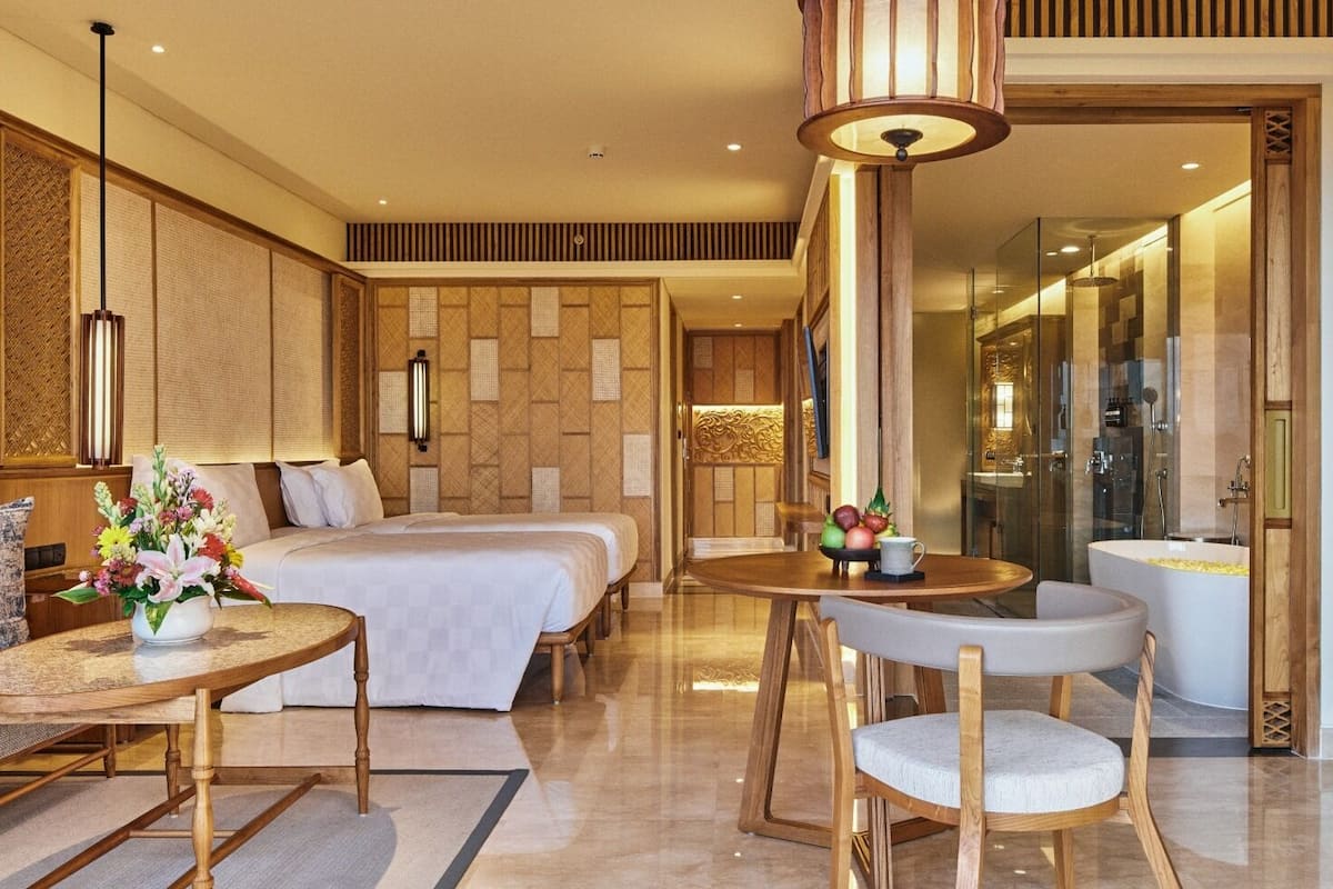 Quarto de hotel com cama, área de estar com mesa e cadeiras, banheiro com banheira e uma janela grande.