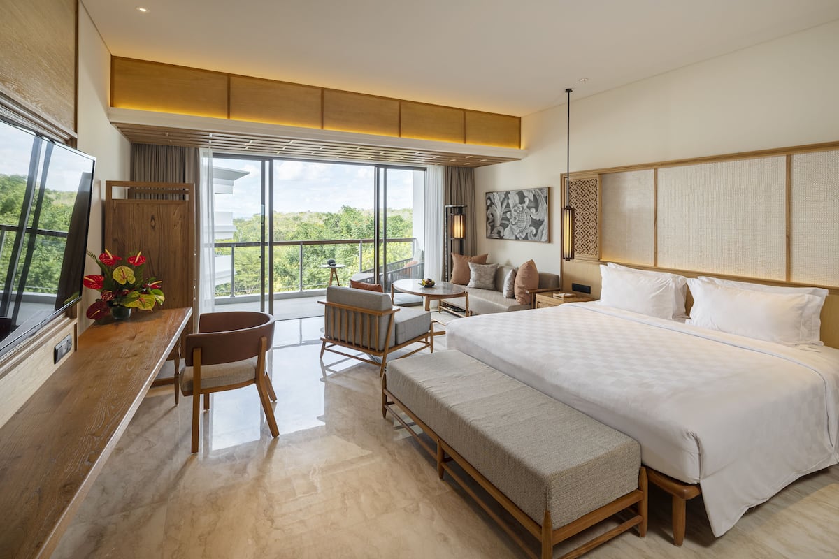 Quarto de hotel moderno com uma cama grande, uma mesa com cadeira, um sofá e uma varanda com vista para o verde.