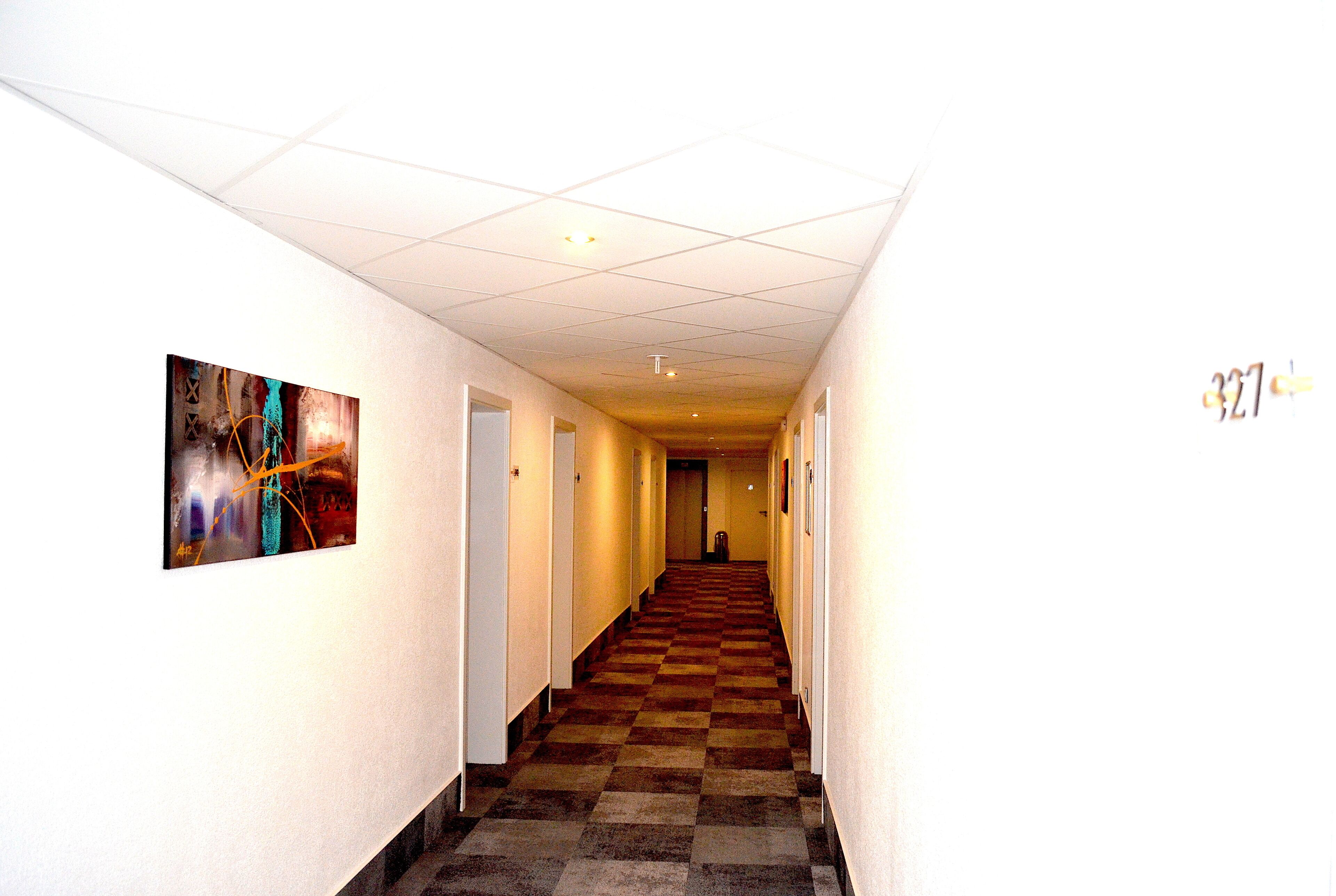 Corredor de hotel com piso xadrez, paredes brancas e uma obra de arte emoldurada à esquerda.