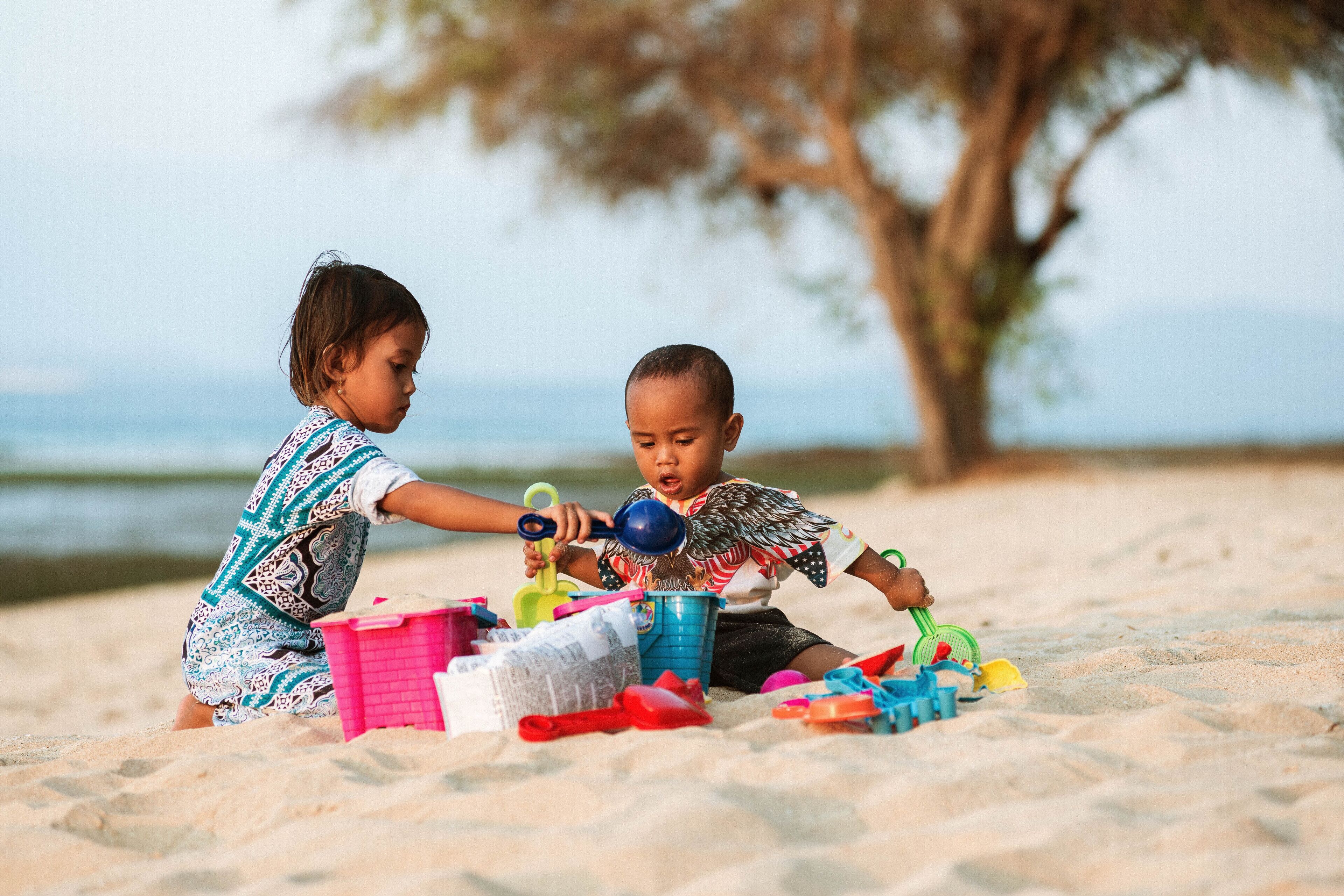 Duas crianças brincando com brinquedos de areia numa praia, com uma árvore e o oceano ao fundo.