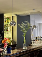 Uma área de bar com um vaso de vidro com flores, uma estante com garrafas de bebidas e luminárias pendentes.