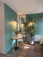 Quarto com paredes verde-água, uma mesa redonda de madeira, duas cadeiras brancas de vime, uma grande planta em vaso e um espelho refletindo o ambiente.