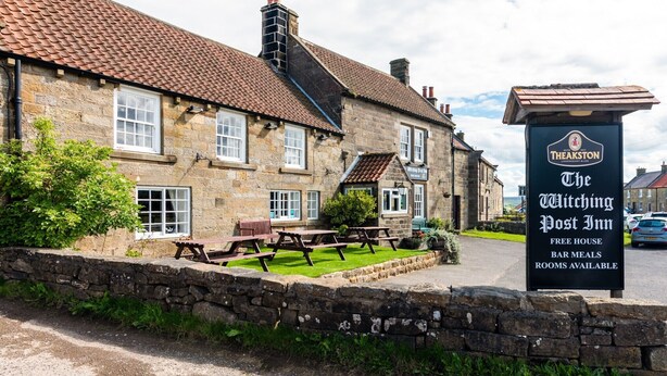 O The Witching Post Inn, um edifício de pedra com design tradicional, exibindo uma placa anunciando bebidas, refeições no bar e quartos disponíveis.