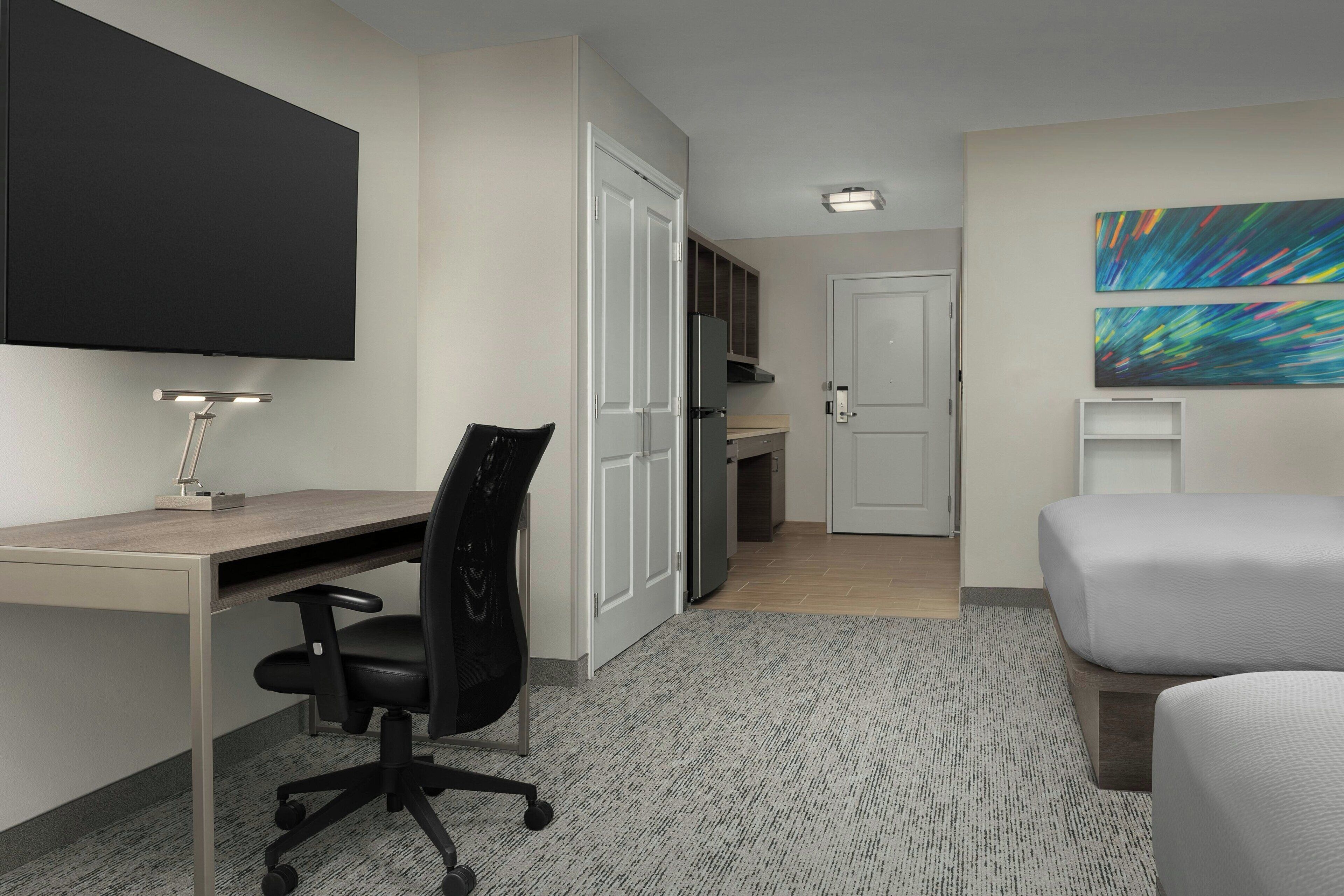Quarto de hotel com escrivaninha, cadeira, cama, obras de arte e porta que dá para uma cozinha compacta.