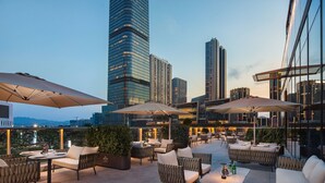 Terraço no topo com área de estar externa, grandes guarda-sóis e vista para o horizonte urbano.