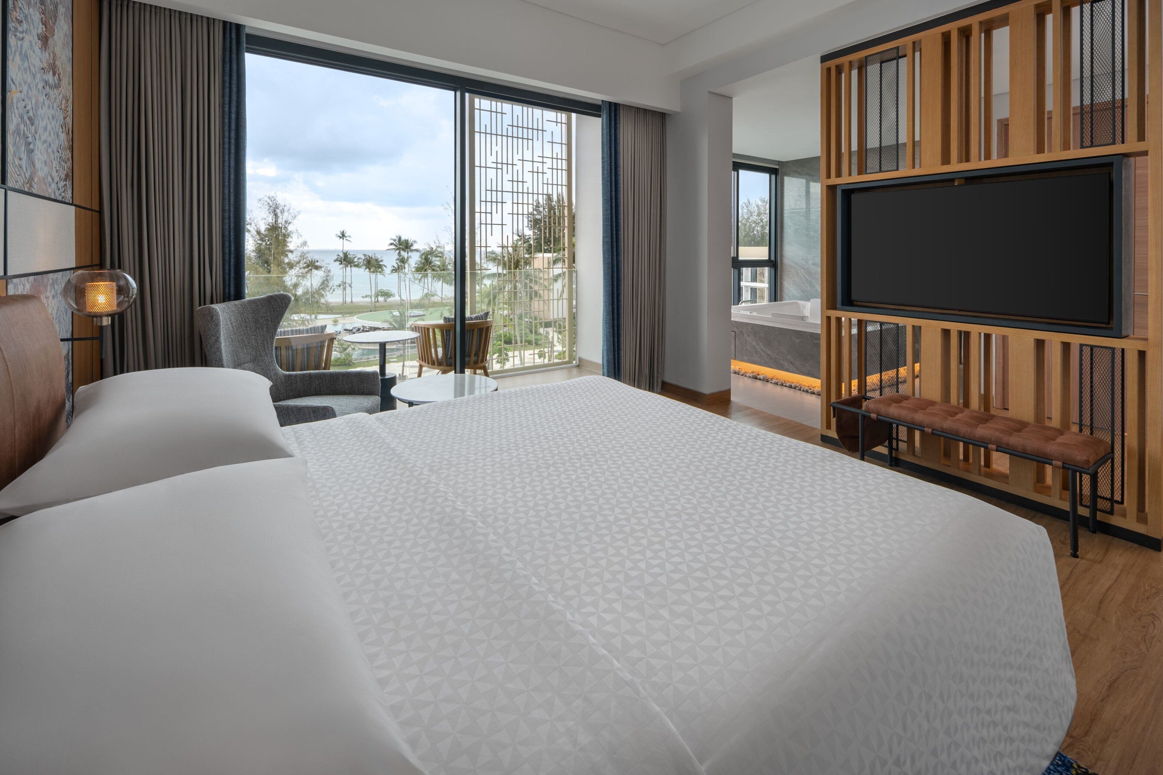Quarto de hotel moderno com uma cama grande, área de estar com mesa e cadeiras, TV fixada em um painel de madeira e varanda com vista para palmeiras.