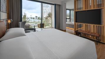 Quarto de hotel moderno com uma cama grande, área de estar com mesa e cadeiras, TV fixada em um painel de madeira e varanda com vista para palmeiras.