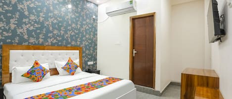 Quarto com cama, cabeceira de madeira, parede com padrão floral, aparelho de ar condicionado, porta de madeira e uma pequena mesa de madeira.