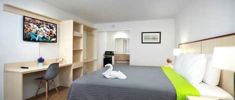 Quarto de hotel com cama, escrivaninha, cadeira, televisão, espelho e mesa de cabeceira.