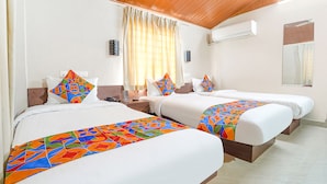 Quarto de hotel com duas camas, cada uma com colchas e almofadas estampadas com padrões geométricos coloridos. Teto de madeira, ar condicionado e janela com cortinas.