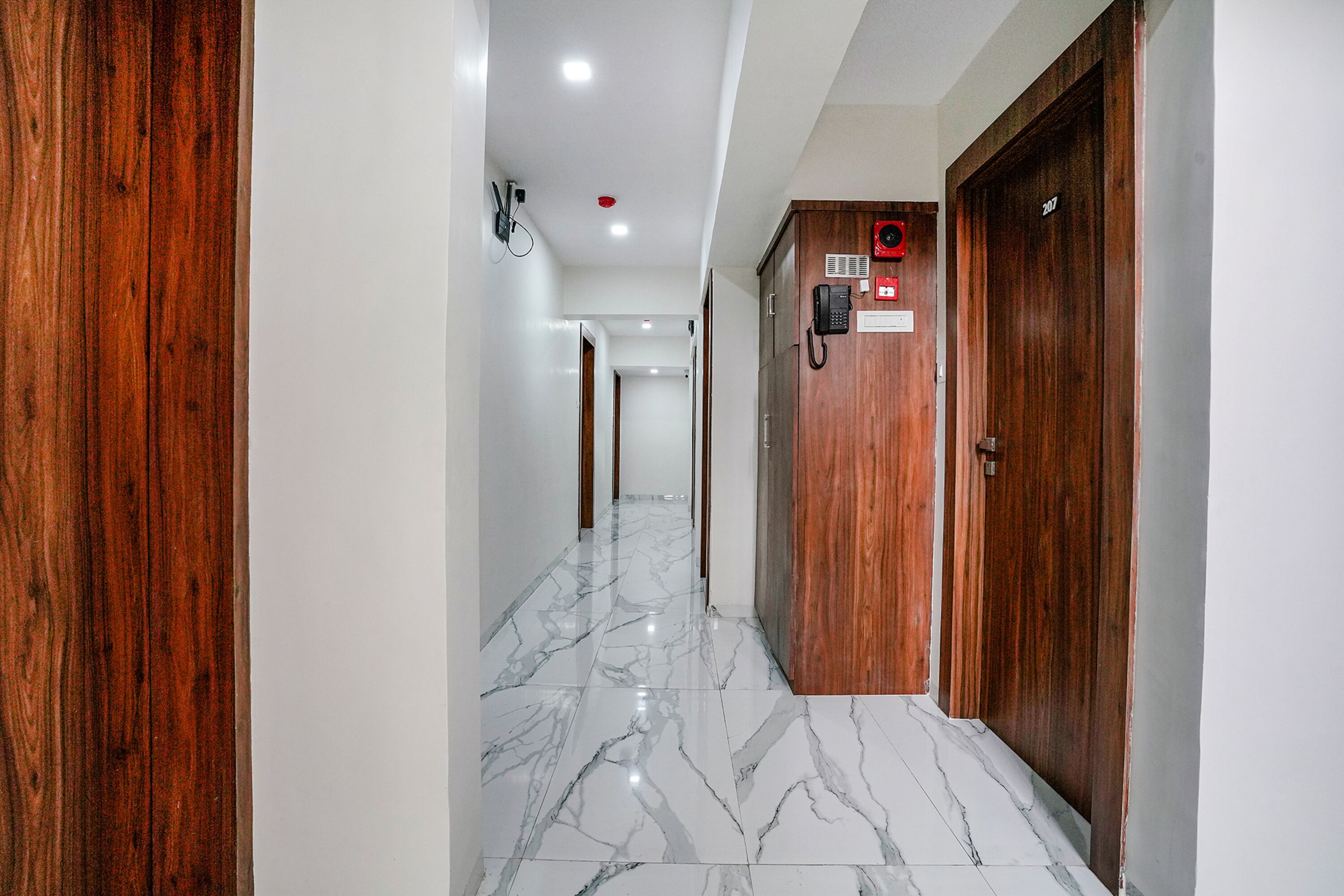 Um corredor com piso de mármore, portas de madeira e um interfone fixado na parede.