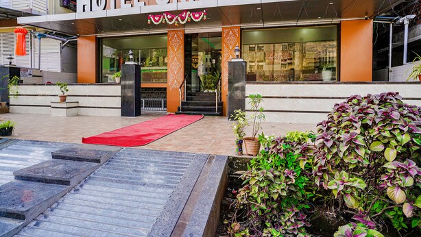 A entrada do Hotel Grand Inn com tapete vermelho, degraus que levam à entrada e plantas em vasos na área.