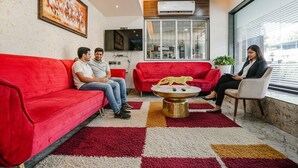 Uma área de espera moderna com sofás vermelhos, uma mesa de centro com um objeto decorativo e uma grande janela com persianas.