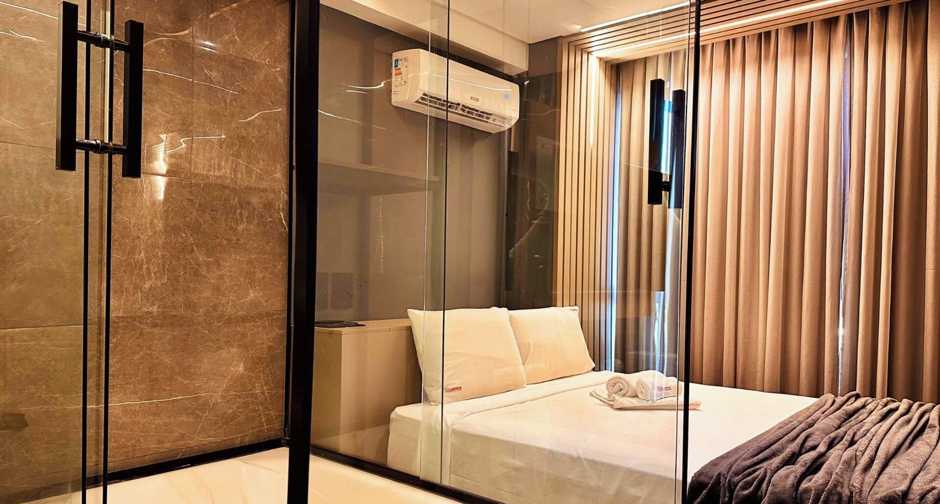 Quarto de hotel moderno com divisória de vidro, cama com roupa de cama branca, manta marrom e ar condicionado fixado na parede.