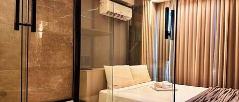 Quarto de hotel moderno com divisória de vidro, cama com roupa de cama branca, manta marrom e ar condicionado fixado na parede.