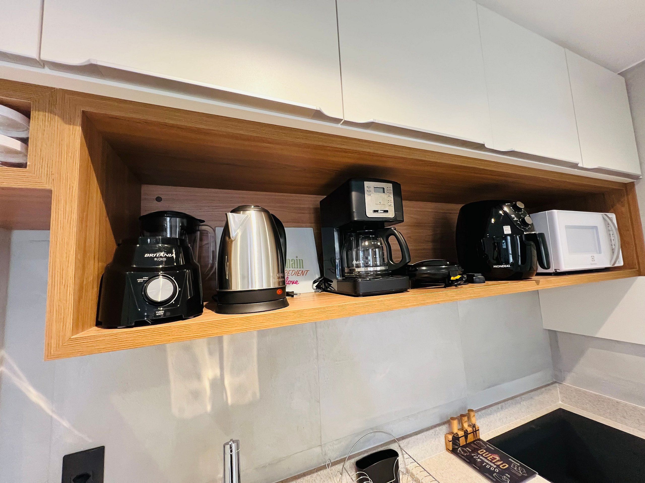 Uma cozinha com prateleira de madeira contendo uma cafeteira, chaleira, micro-ondas e torradeira.