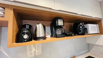 Uma cozinha com prateleira de madeira contendo uma cafeteira, chaleira, micro-ondas e torradeira.
