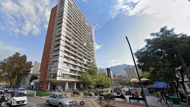 Um edifício alto e moderno, com vários andares, cercado por árvores e uma rua movimentada.