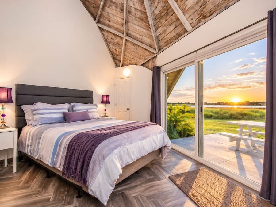 Quarto com cama grande, mesas de cabeceira, abajur e vista para o pôr do sol através de uma porta de correr.