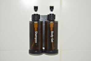 Duas garrafas transparentes com bombas, identificadas como 'Shampoo' e 'Gel de Banho', fixadas numa parede.