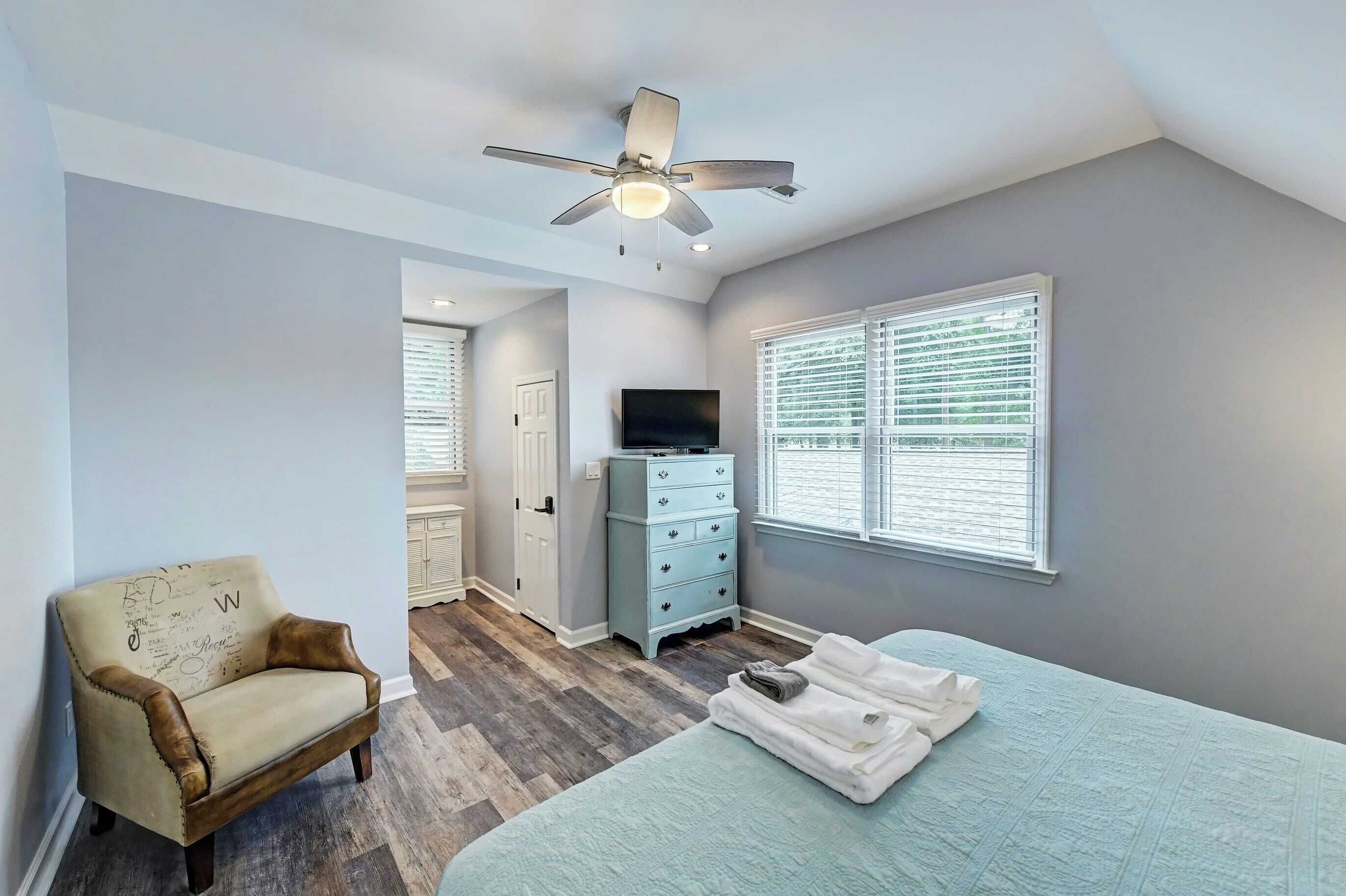 Um quarto com cama, cadeira, cômoda, ventilador de teto e janela.