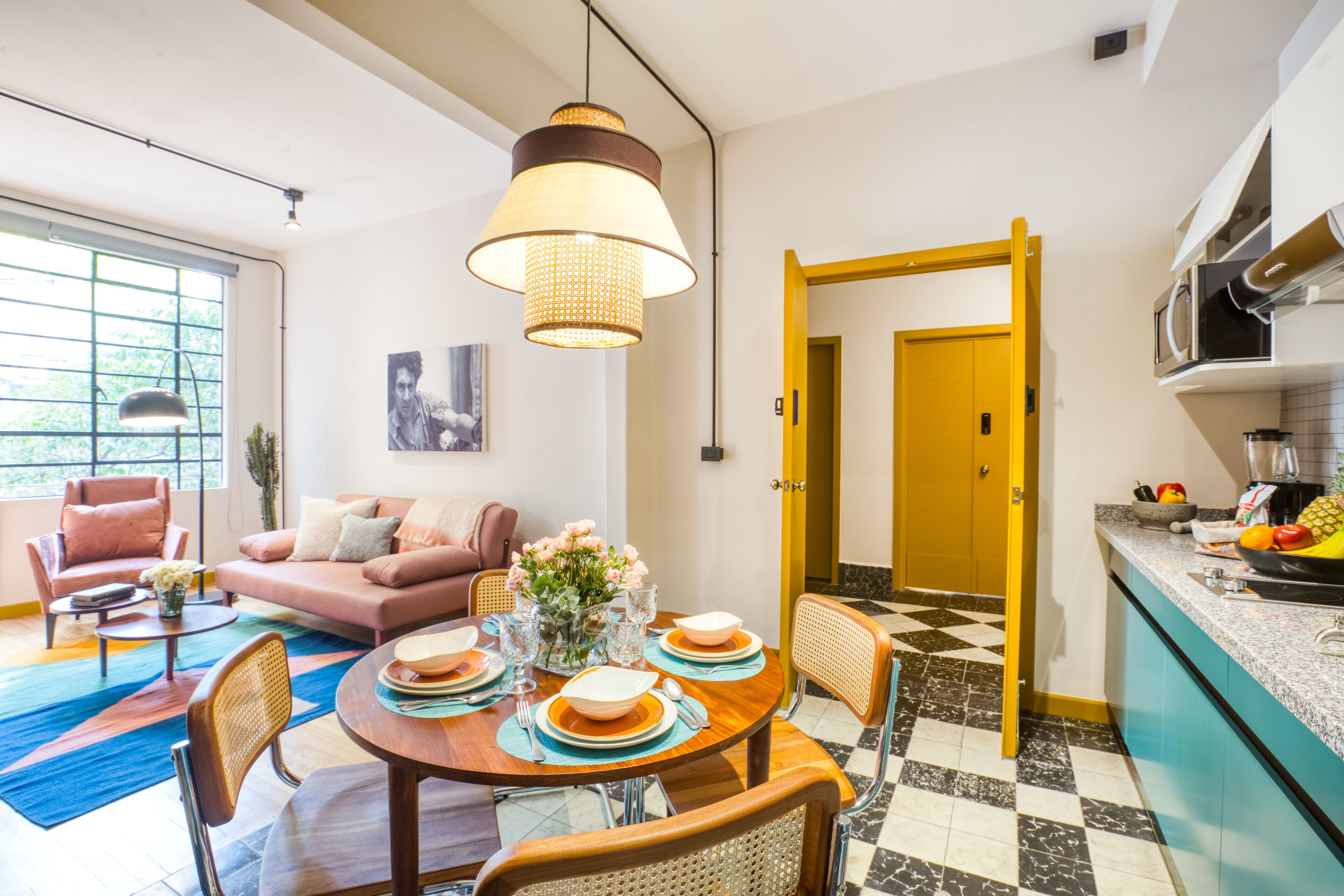 Uma sala de estar moderna com sofá, mesa de centro e mesa de jantar posta com pratos e copos. Uma luminária pendente grande está suspensa acima da mesa.