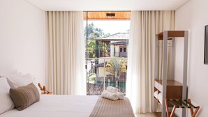Quarto de hotel com uma cama grande, um banquinho de madeira e vista para um edifício pela janela.