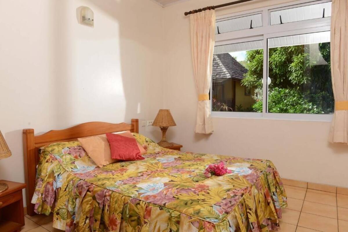 Quarto com colcha floral, cabeceira de madeira, luminária de cabeceira e janela com cortinas.