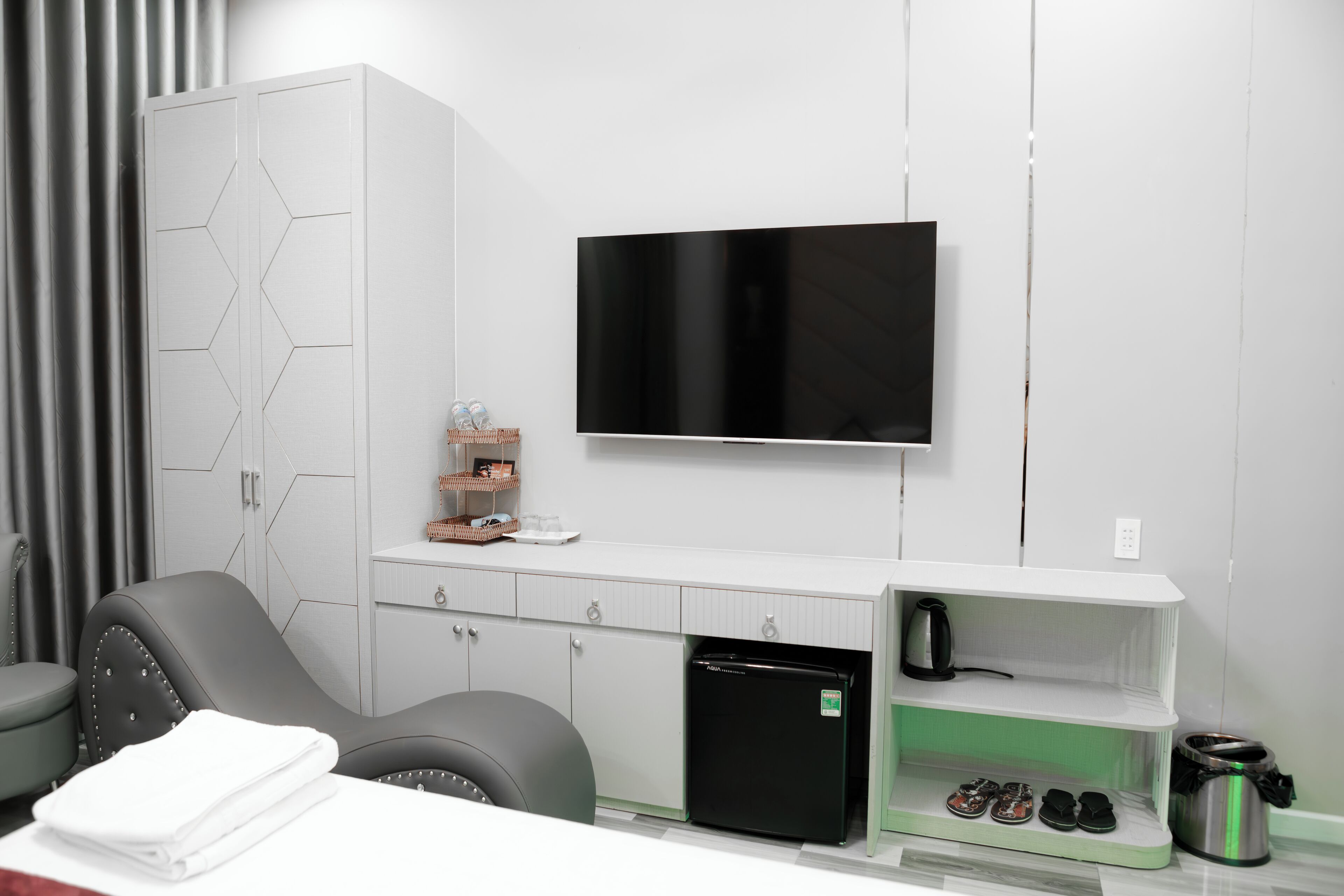 Quarto moderno e bem iluminado, com televisão de tela plana, pequena geladeira, máquina de café e chinelos organizados.