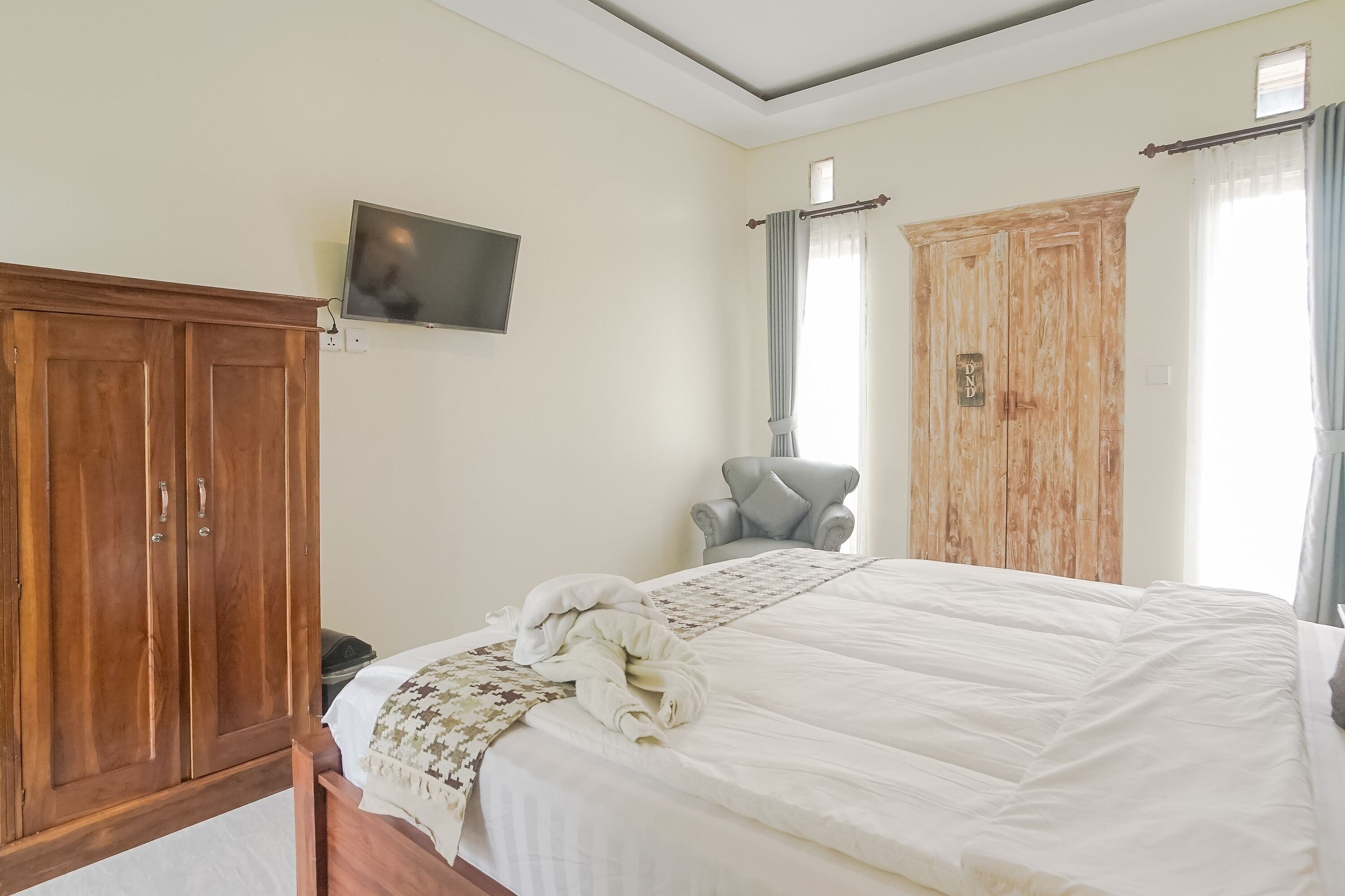 Quarto com cama grande, guarda-roupa de madeira, televisão na parede e uma cadeira.