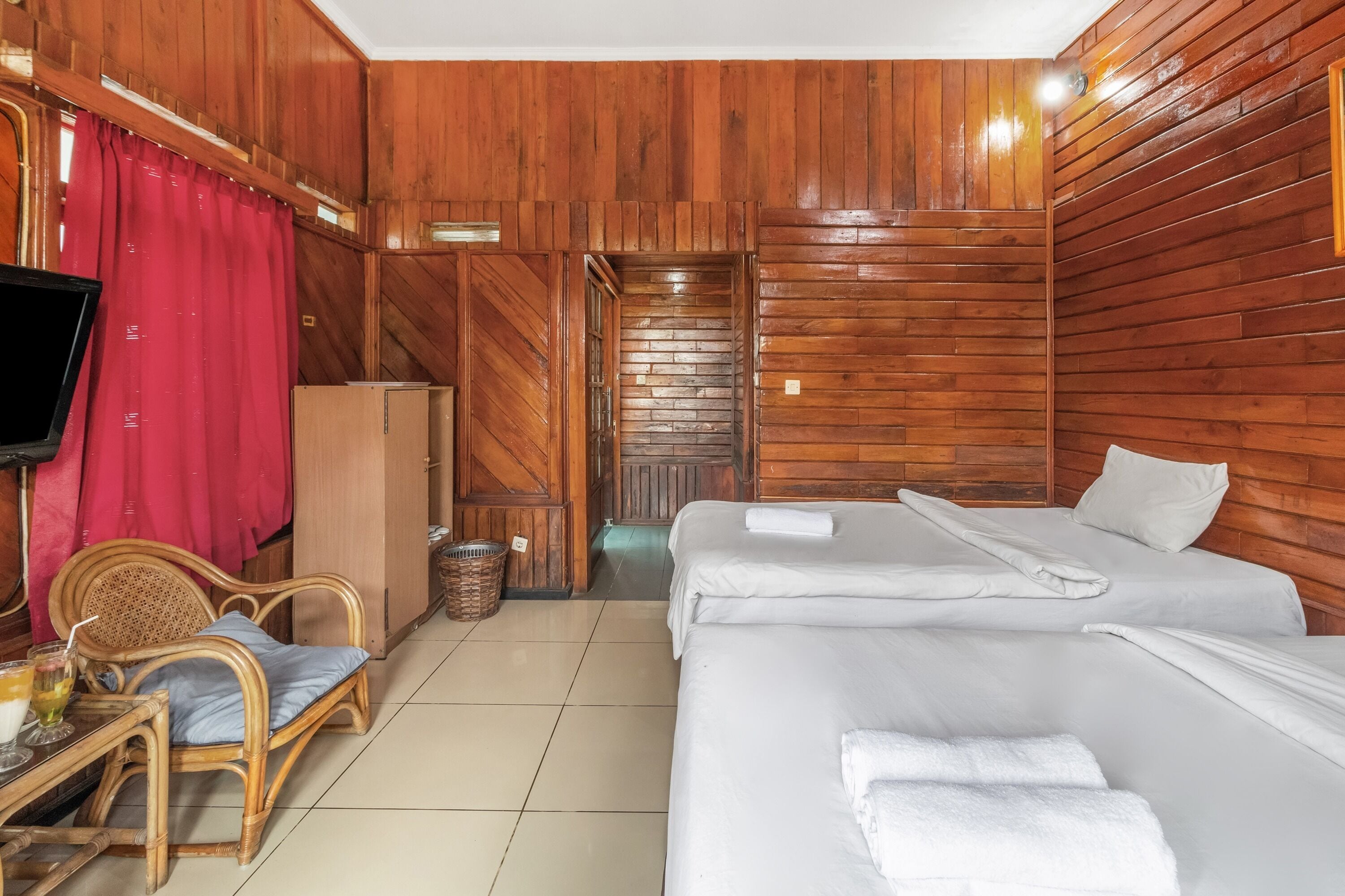 Quarto de hotel com painéis de madeira, duas camas com lençóis brancos, uma cadeira de madeira, uma televisão e uma cortina vermelha.