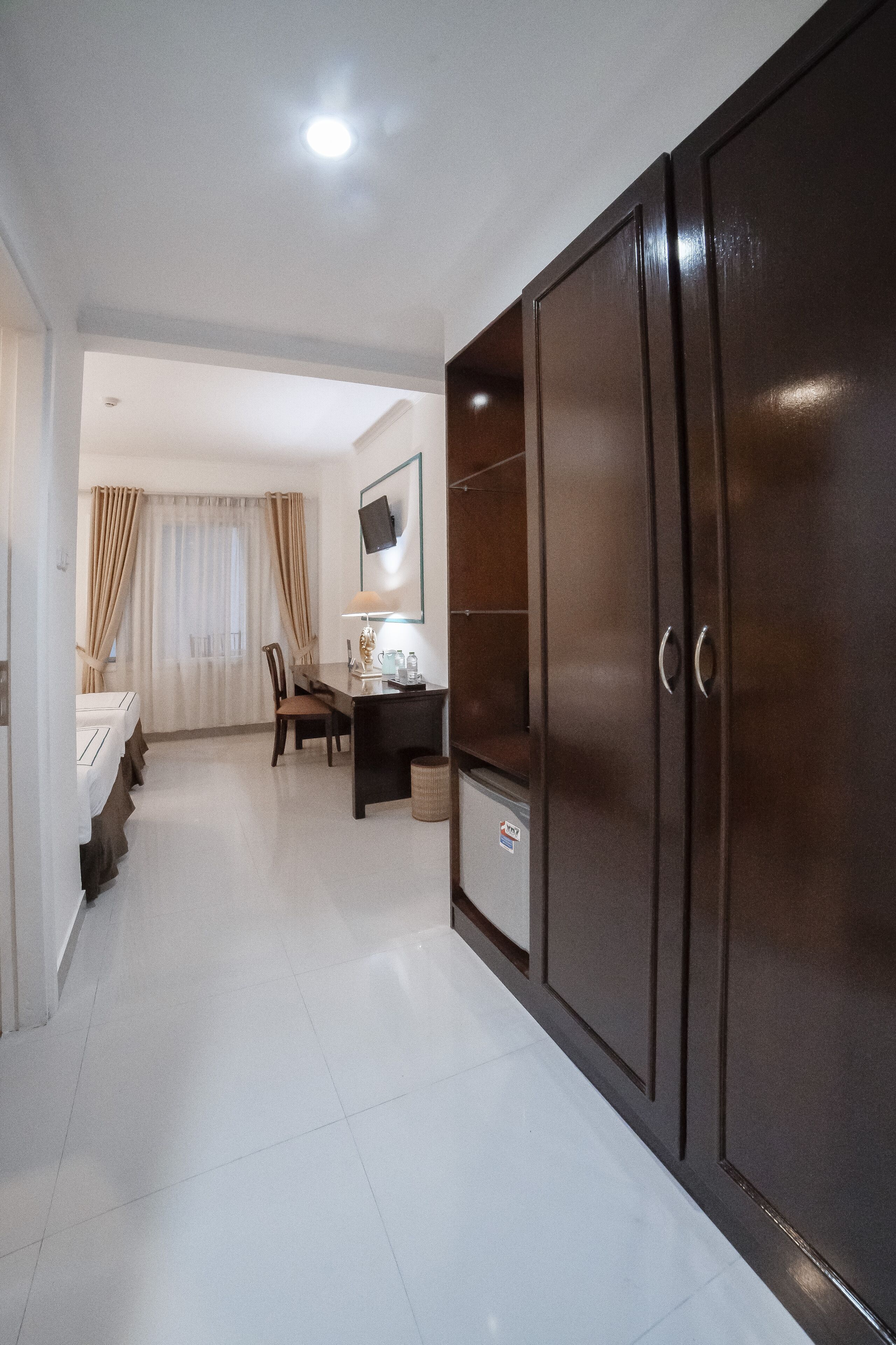 Quarto de hotel com um guarda-roupa grande, uma escrivaninha com cadeira, uma cama e uma televisão na parede.
