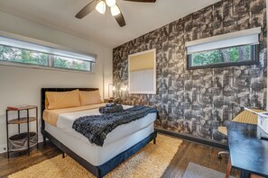 Quarto com cama, escrivaninha, ventilador de teto e grandes janelas.