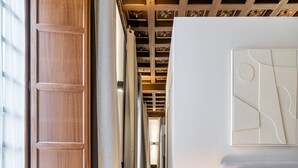 Quarto de hotel com piso de madeira, cama com roupa de cama branca, mesa de cabeceira com abajur e vista para uma área de refeições através de uma porta de vidro.